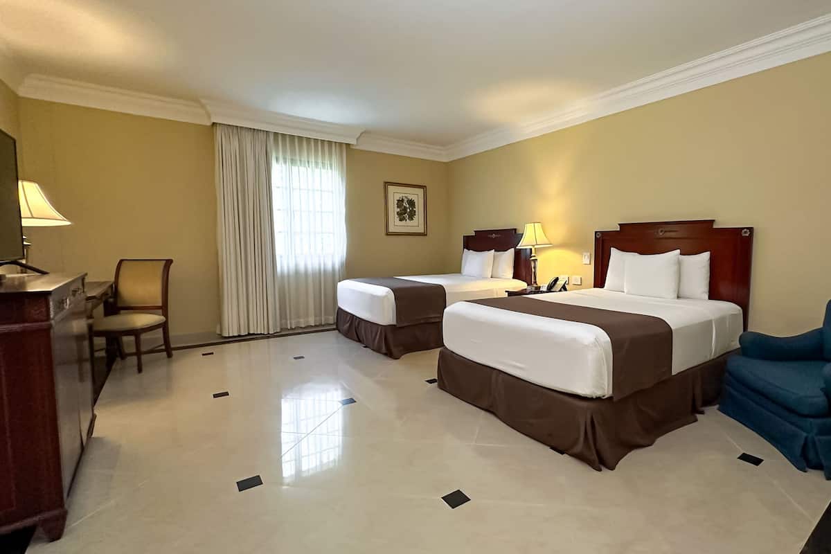 Quarto de hotel com duas camas, uma escrivaninha, uma cadeira e uma poltrona azul.