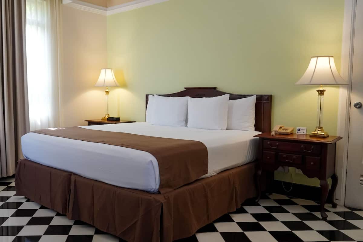Quarto de hotel com piso xadrez, cama com roupa de cama branca e marrom, duas luminárias de cabeceira, uma mesinha de cabeceira de madeira e uma escrivaninha com telefone.