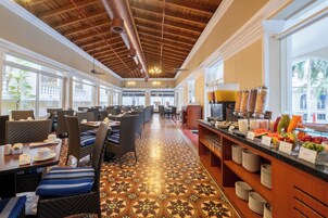 Restaurante com tetos altos, ladrilhos estampados no chão e uma área de buffet com frutas e bebidas.