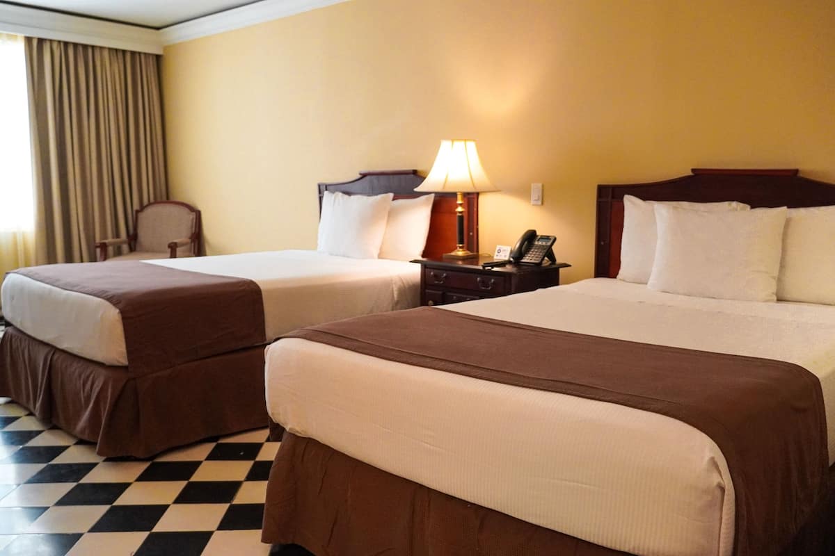 Duas camas de casal com colchas marrons, uma mesa de cabeceira com abajur e uma cadeira em quarto de hotel com chão quadriculado.