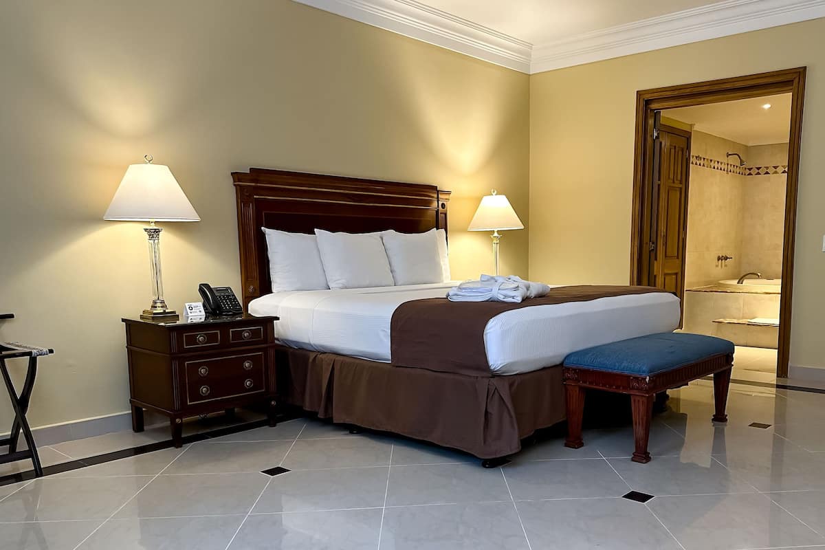 Quarto de hotel com uma cama grande, duas luminárias de cabeceira, uma mesa de cabeceira de madeira, uma escrivaninha com telefone e um banheiro visível por uma porta aberta.