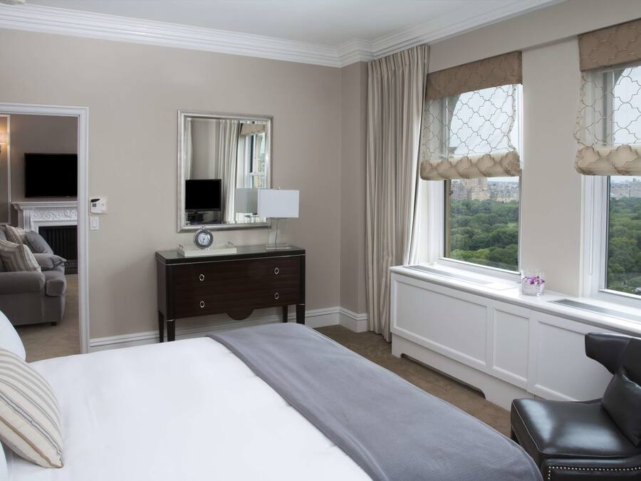 Quarto de hotel com uma cama grande, cômoda, cadeira, televisão e janela com vista para árvores.
