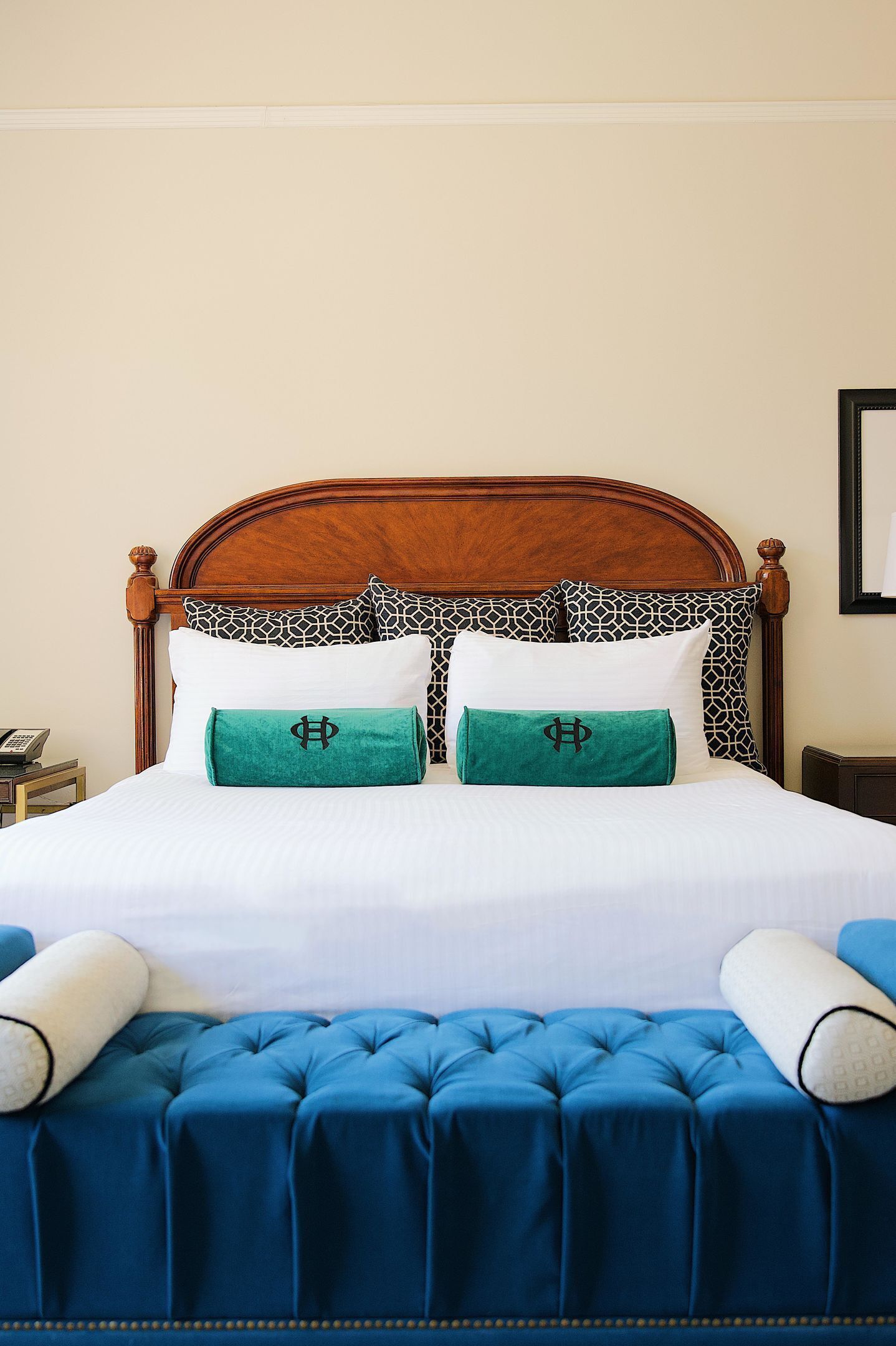 Cama bem arrumada com cabeceira de madeira, duas almofadas verdes com logotipo e cabeceira estofada azul.