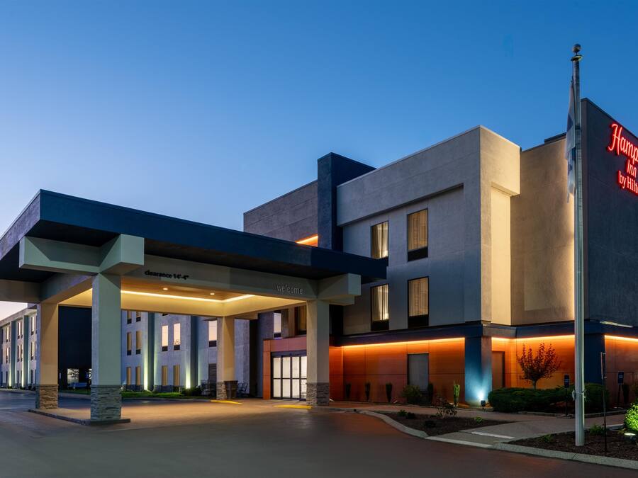 Um hotel moderno com entrada desimpedida, placa indicando ser um Hampton Inn by Hilton, e área coberta com um letreiro de 'Welcome'.