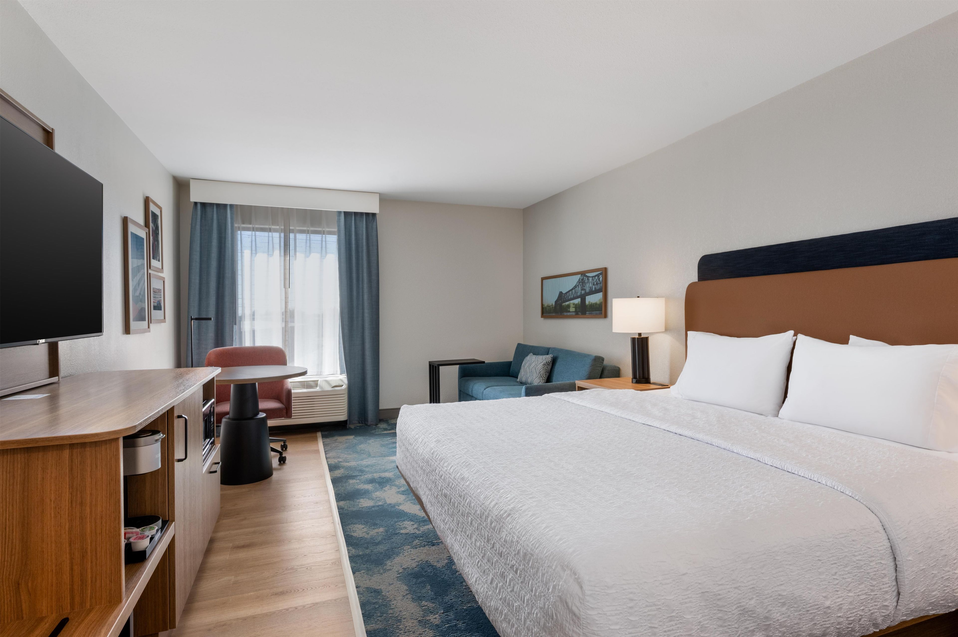 Quarto de hotel com cama, escrivaninha, cadeira, televisão, janela com cortinas, um quadro na parede e abajur.