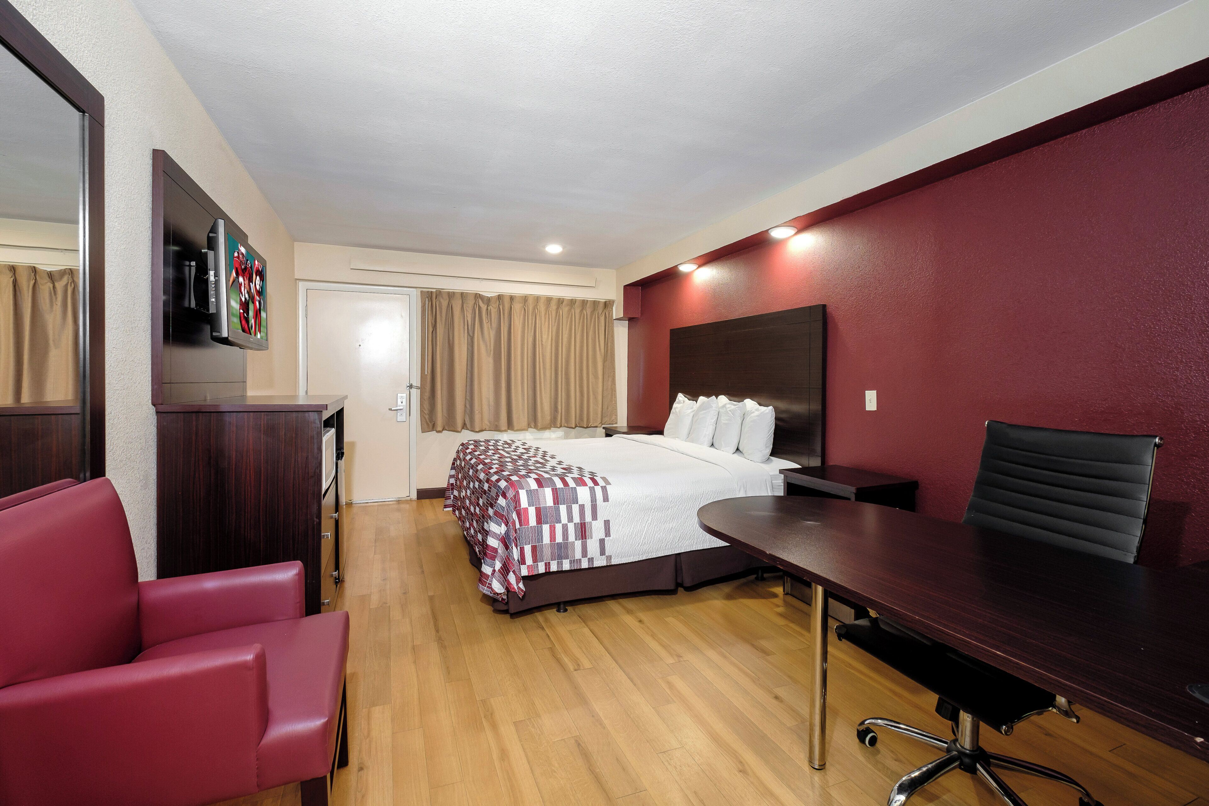 Quarto de hotel com cama, escrivaninha, cadeira, televisão e parede vermelha.