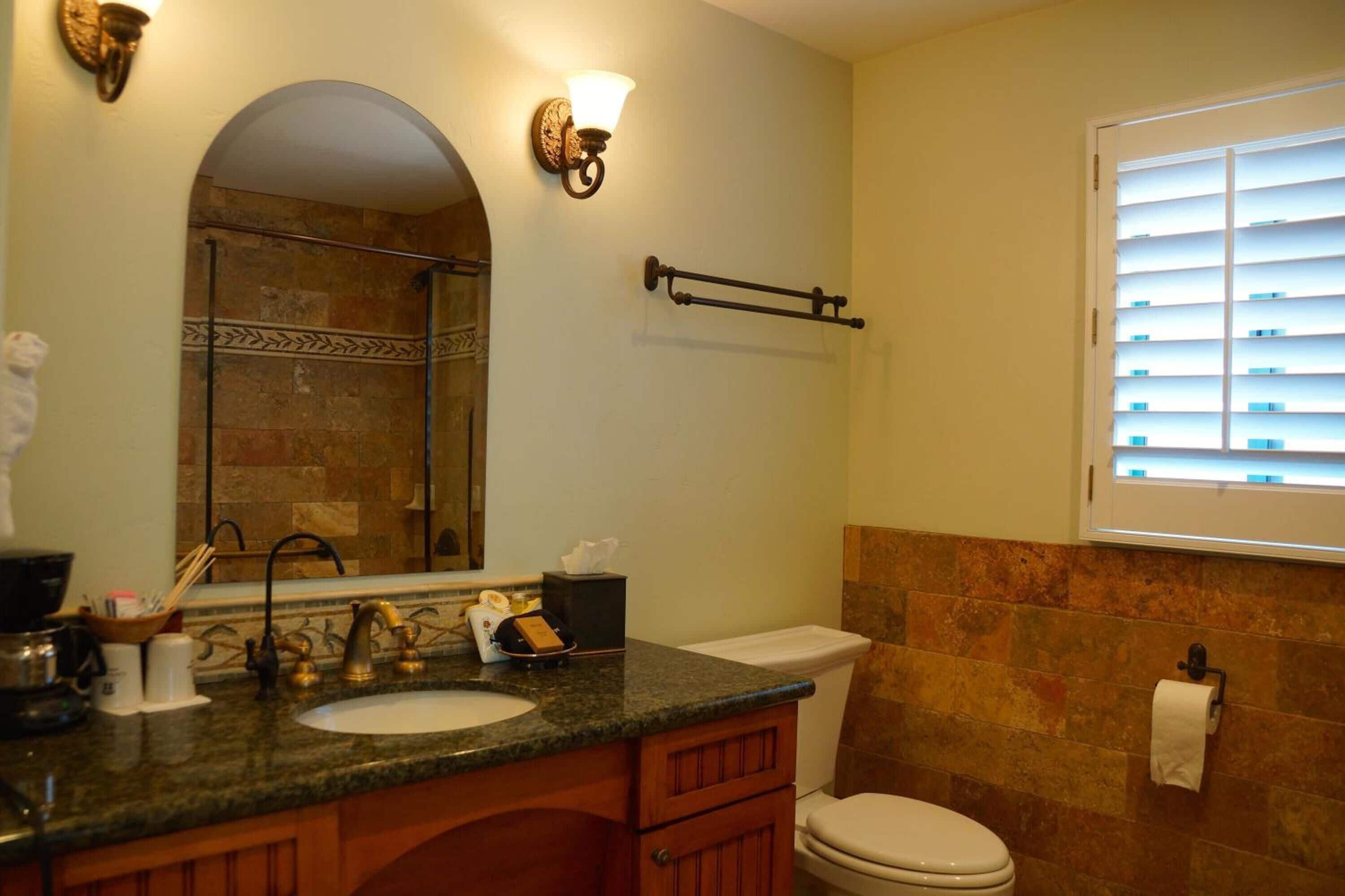 Casa de banho com lavatório bancada em granito, espelho, porta-toalhas, sanita e janela com persianas.