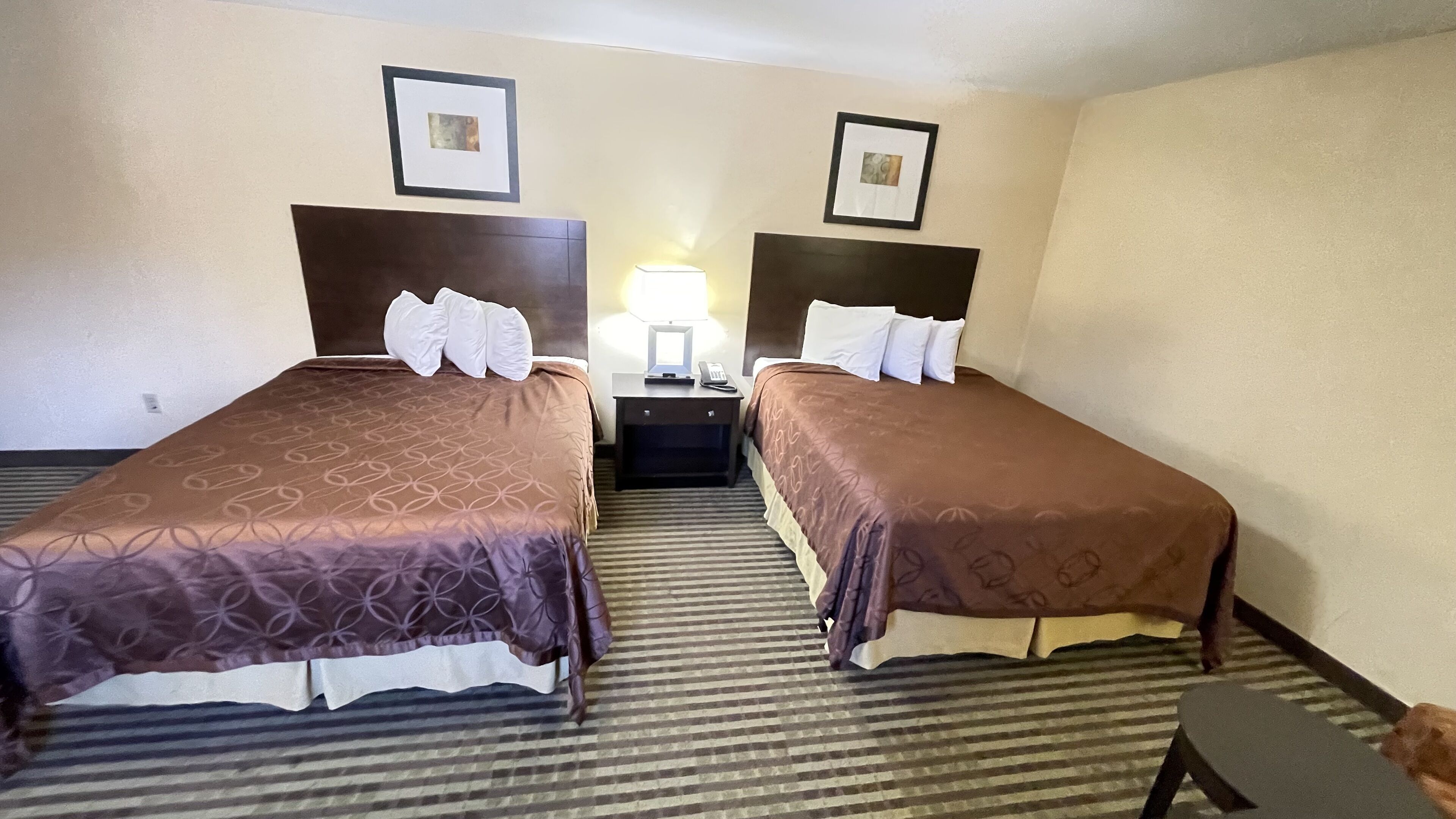 Quarto de hotel com duas camas, uma mesa de cabeceira com abajur e uma pequena mesa.