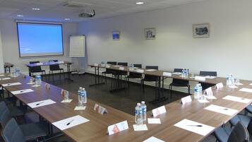 Sala de conferências com mesas compridas, cadeiras, placas de identificação, garrafas de água, tela de projetor e quadro branco.