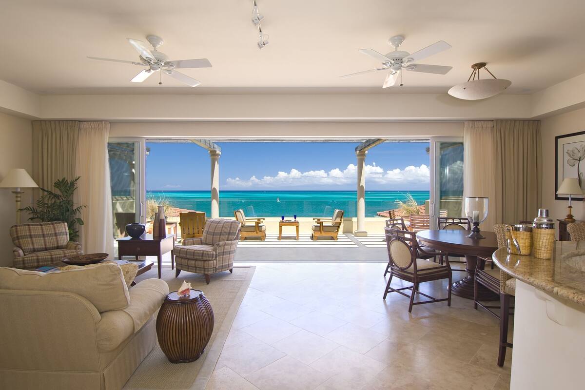 Uma sala de estar ampla com uma grande janela com vista para uma praia, mobiliada com um sofá, poltronas e uma mesa de jantar.