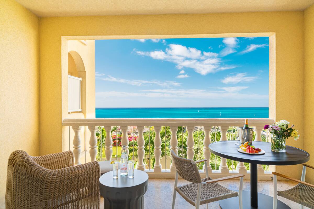 Varanda com móveis de vime, mesa redonda com uma garrafa, copos e uma travessa de fruta, com vista para o oceano.