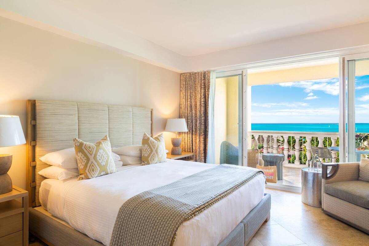Quarto de hotel com cama grande, duas mesas de cabeceira com abajures, varanda com vista para o oceano e um espaço de estar com uma cadeira e uma mesa pequena.