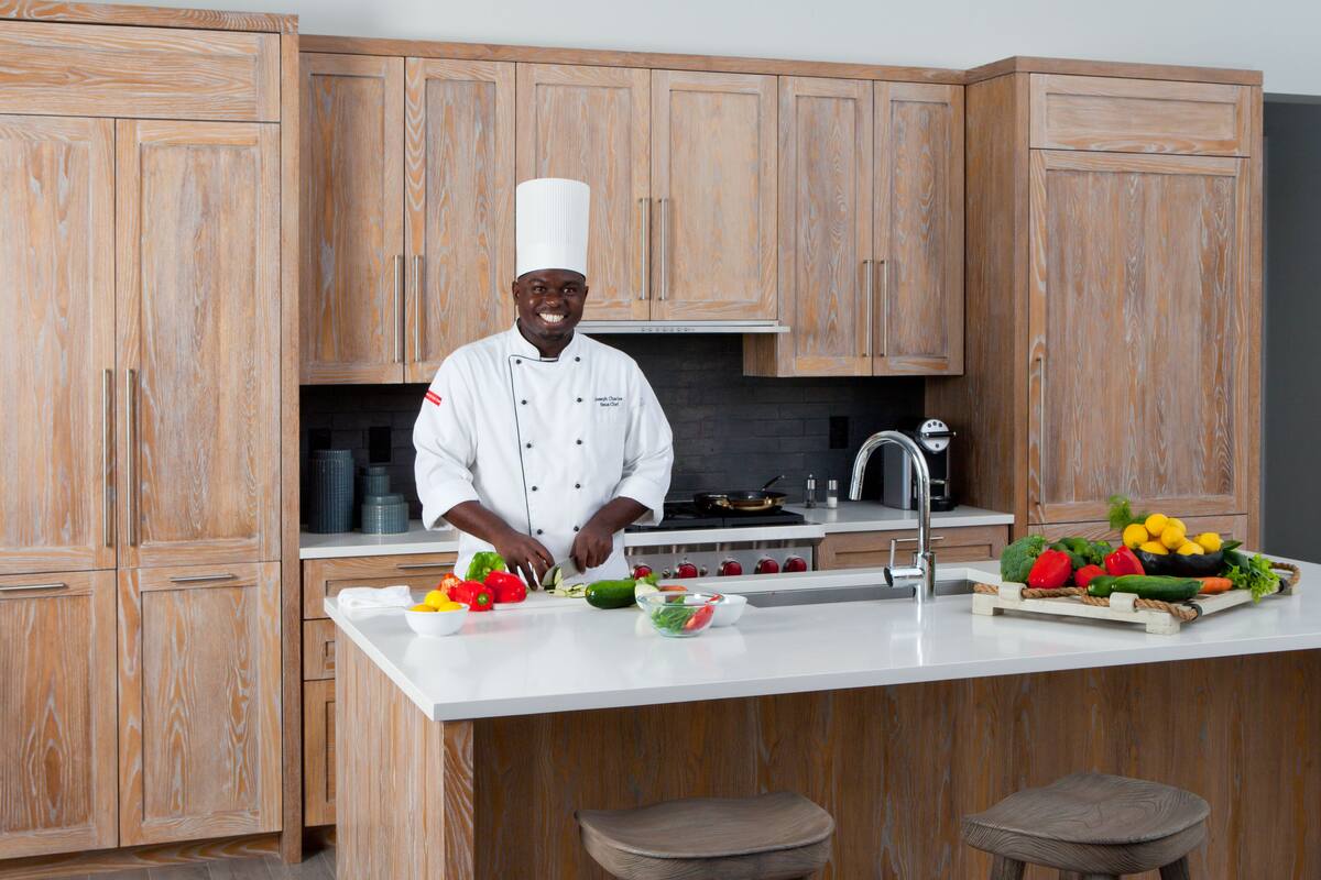 Um chef com uniforme branco prepara vegetais em uma cozinha moderna com armários de madeira e bancada branca.
