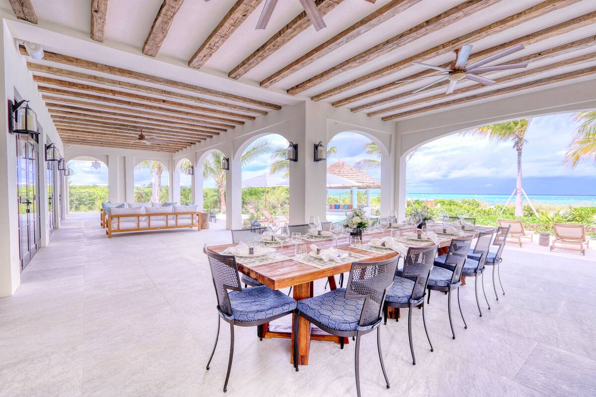 Uma área de refeições ao ar livre espaçosa, com mesa e cadeiras de madeira, janelas amplas e vista para o oceano.