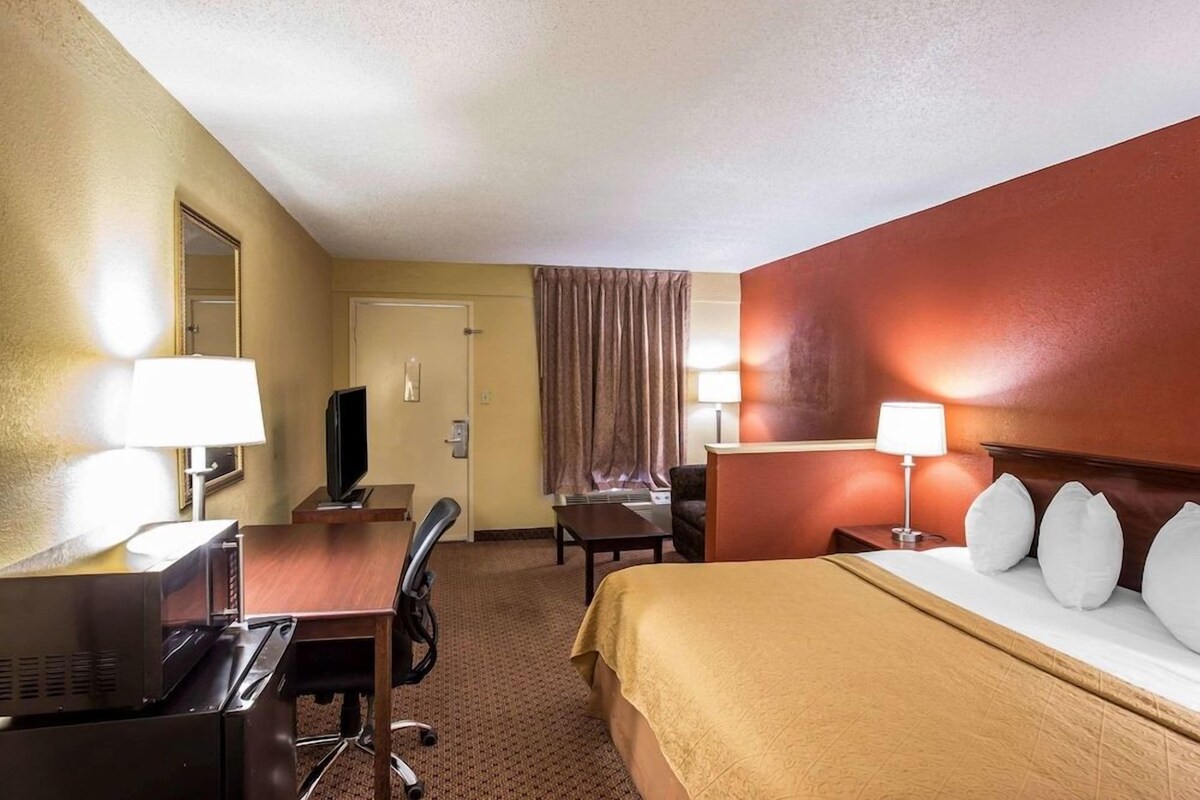 Quarto de hotel com cama, escrivaninha, cadeira, televisão e luminárias.