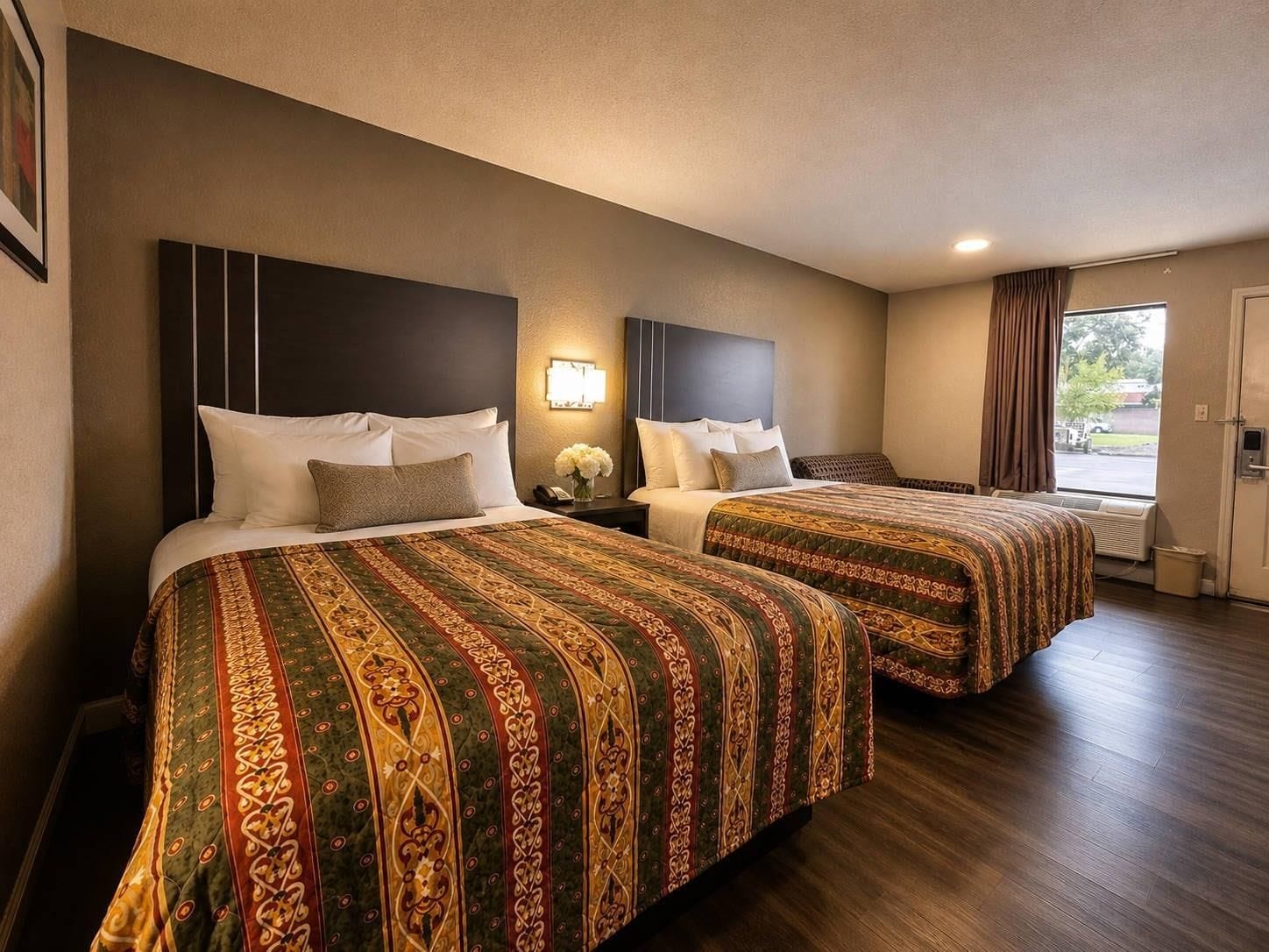 Quarto de hotel com duas camas, uma mesa de cabeceira, uma luminária e uma janela com cortinas.