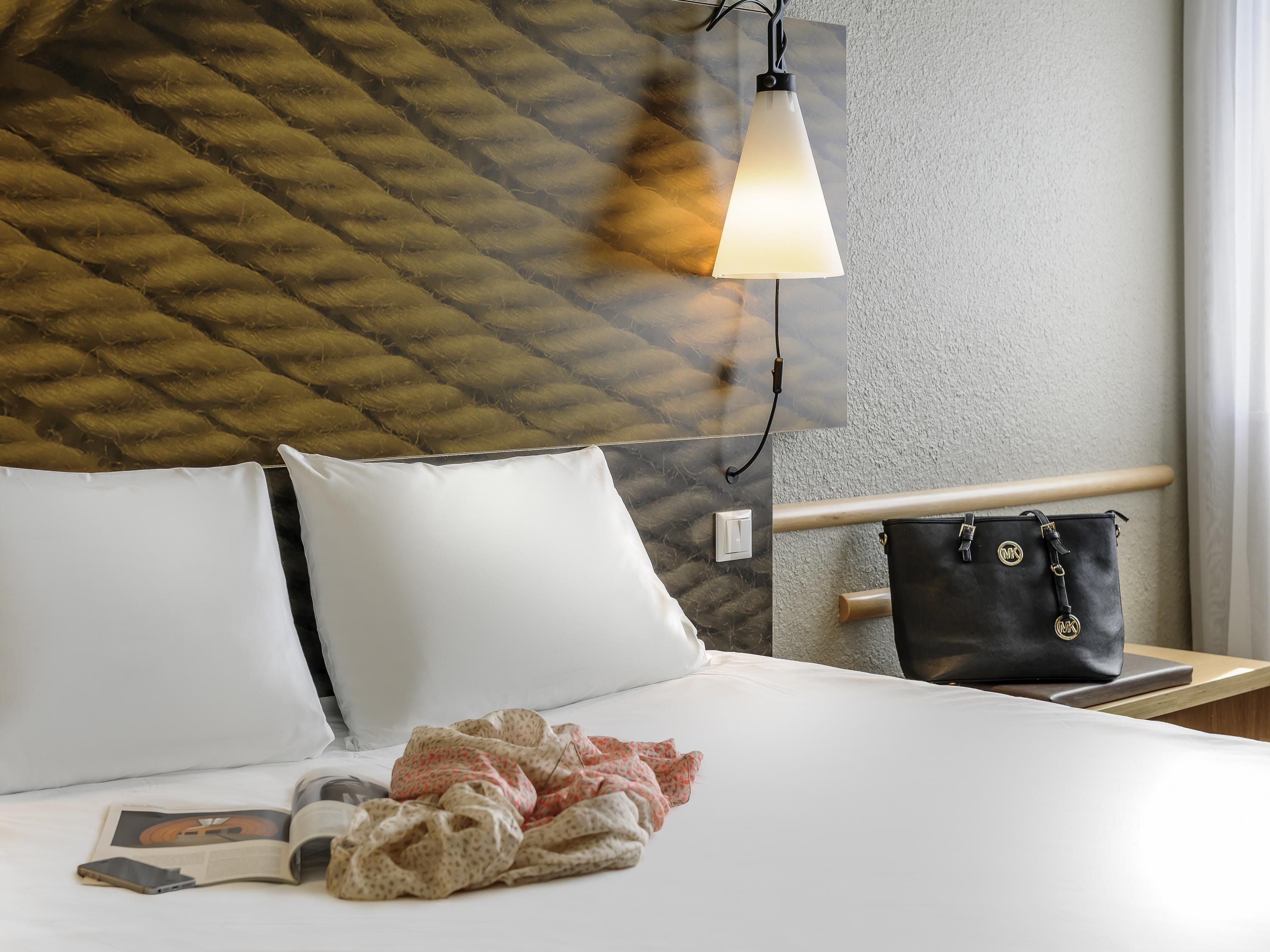 Quarto de hotel com cama, almofadas, abajur, uma bolsa e uma revista sobre a cama.