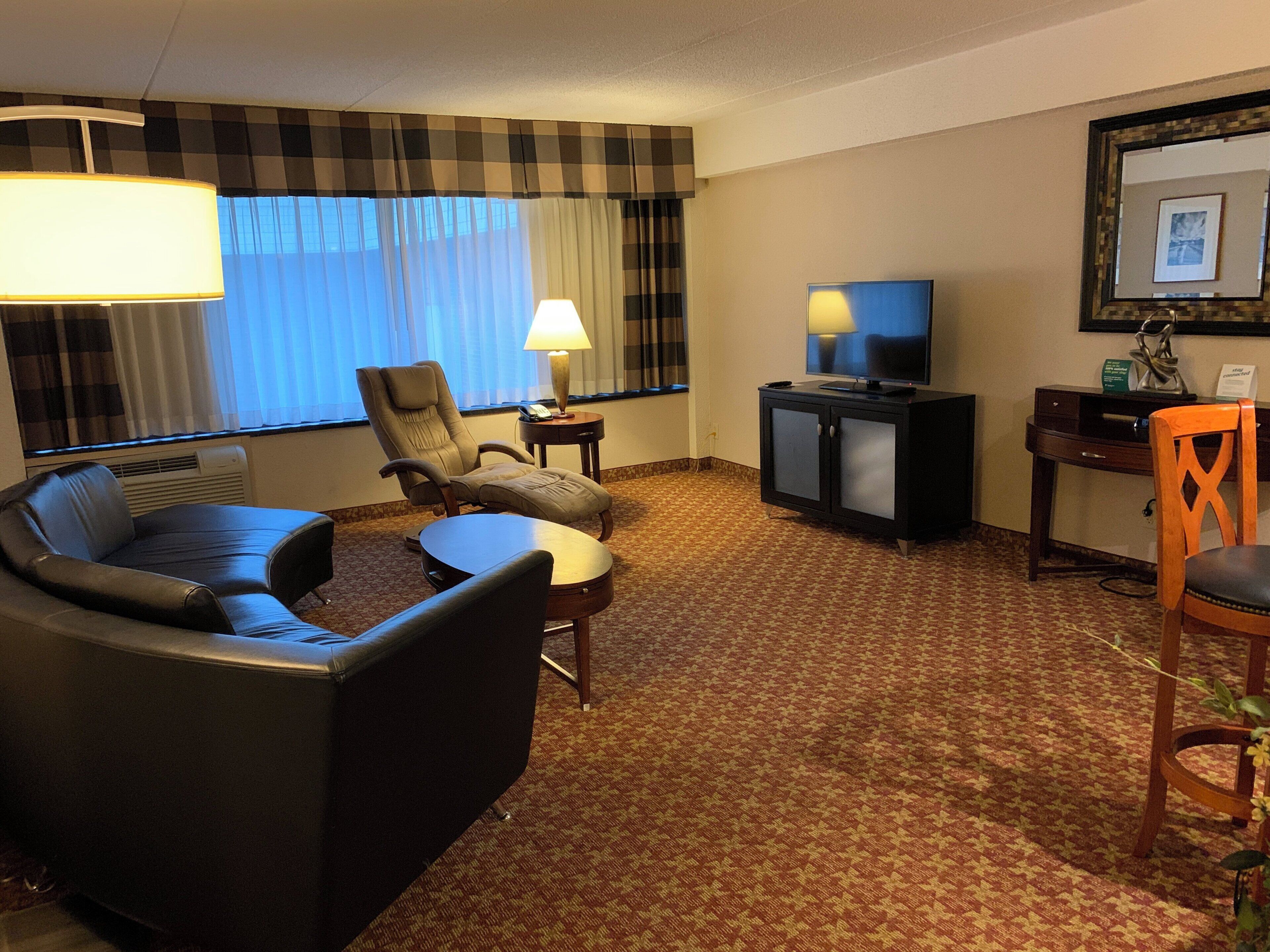 Quarto de hotel com sofá, poltrona, mesa pequena, televisão e escrivaninha com luminária.