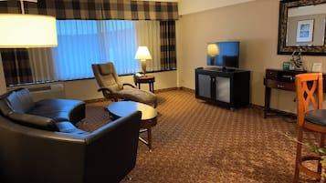 Quarto de hotel com sofá, poltrona, mesa pequena, televisão e escrivaninha com luminária.