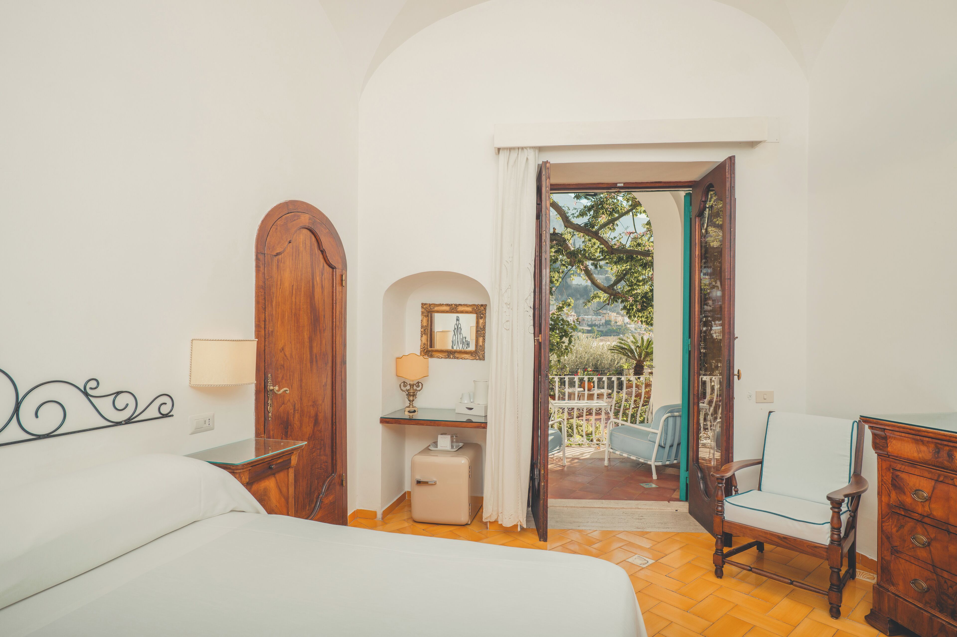 Quarto com cama, escrivaninha, cadeira e guarda-roupa. Há uma porta que dá para área externa com vista para árvores e uma cadeira azul.
