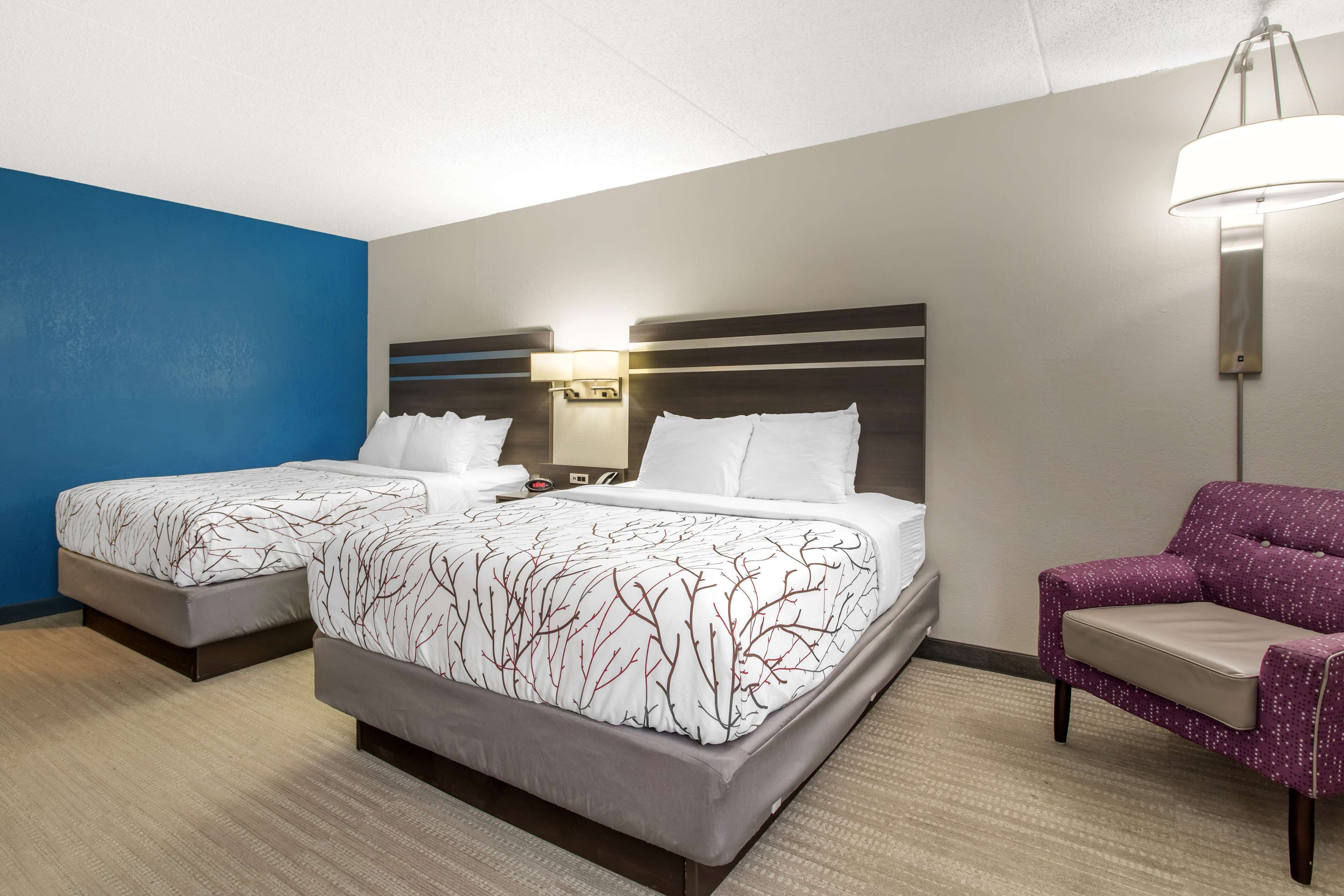Quarto de hotel com duas camas, uma mesa de cabeceira, um abajur e uma poltrona roxa.