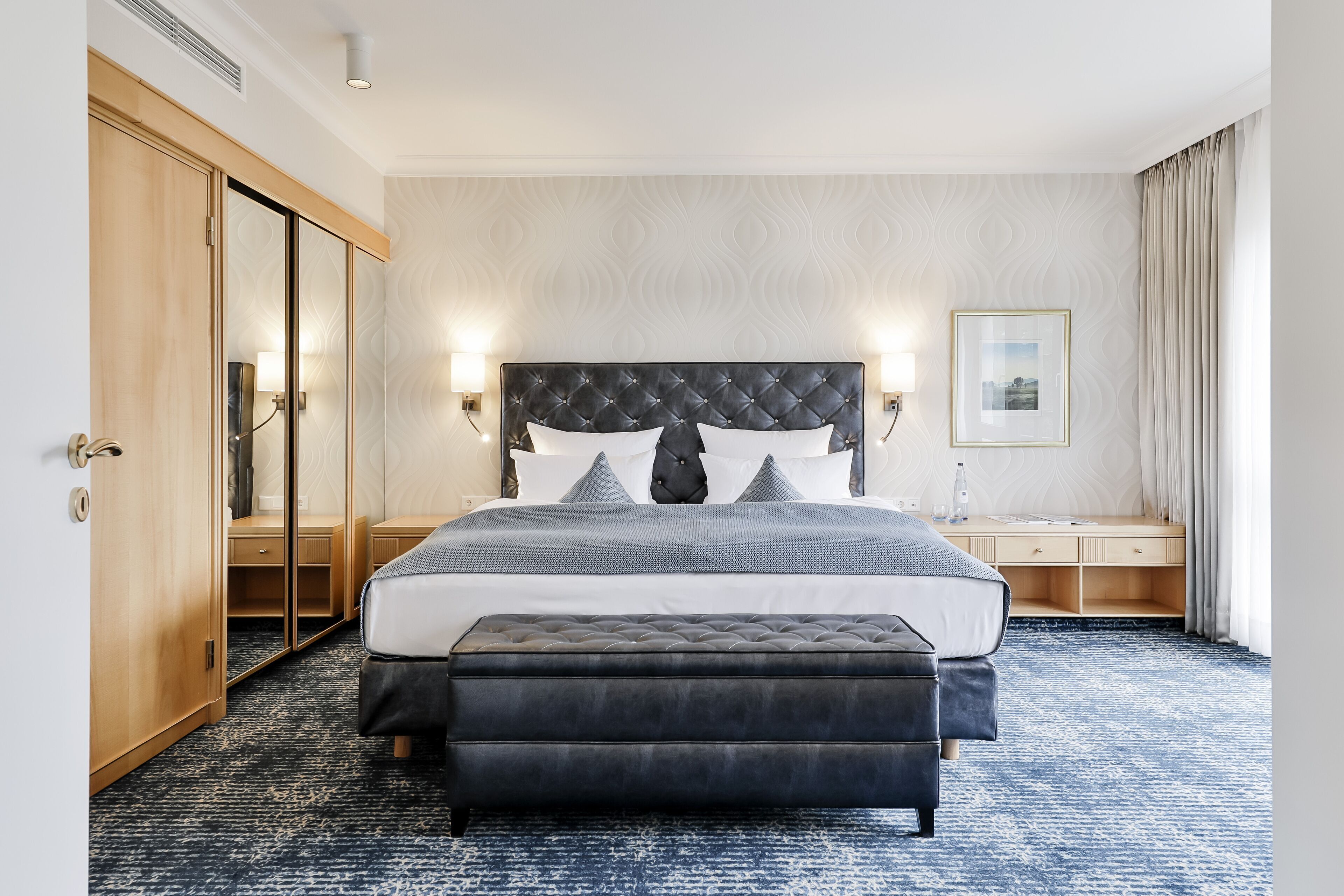 Quarto de hotel com uma cama grande, um guarda-roupa, uma escrivaninha e uma janela com cortinas.
