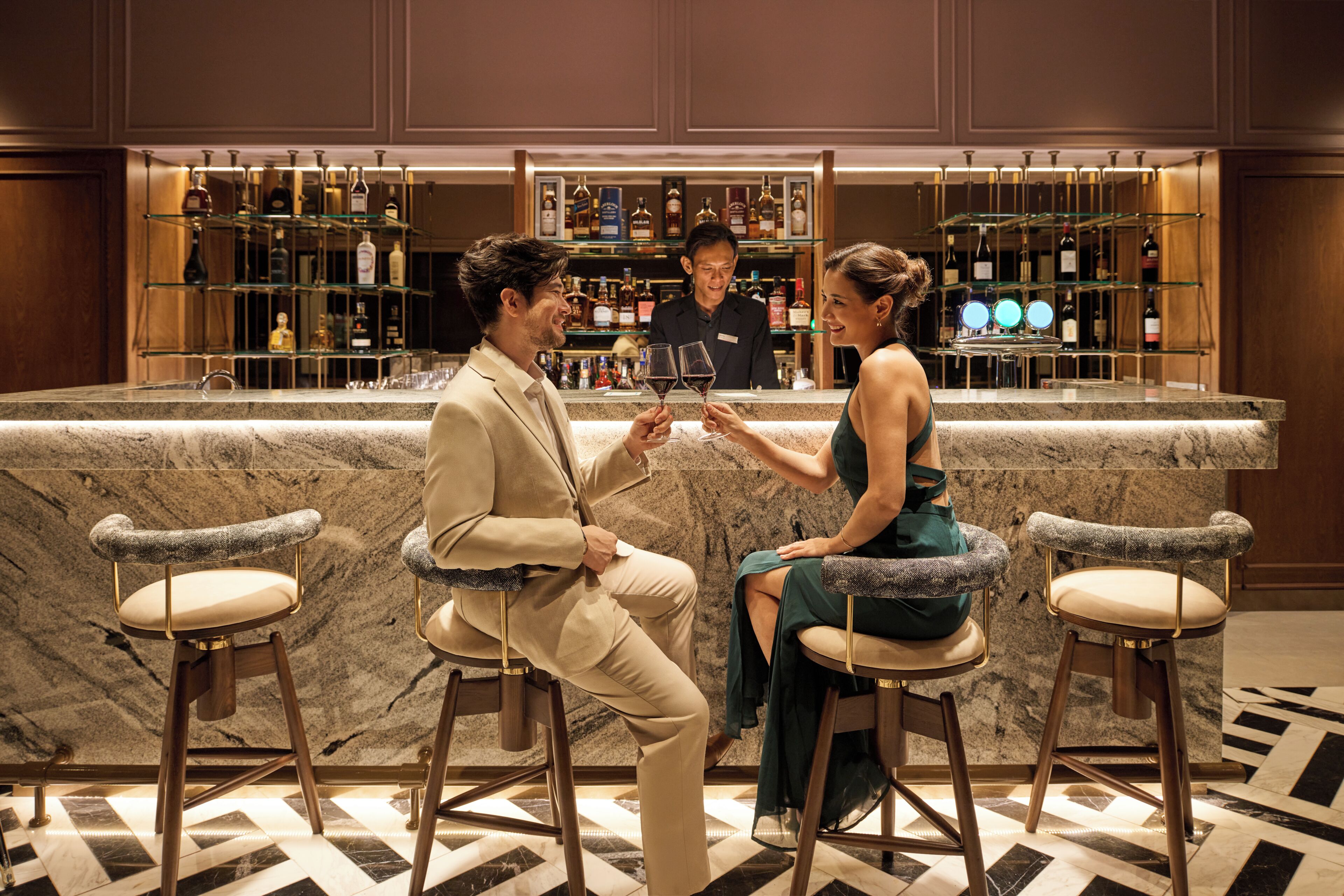 Uma área de bar com bancada de mármore, várias prateleiras cheias de garrafas e dois bancos.