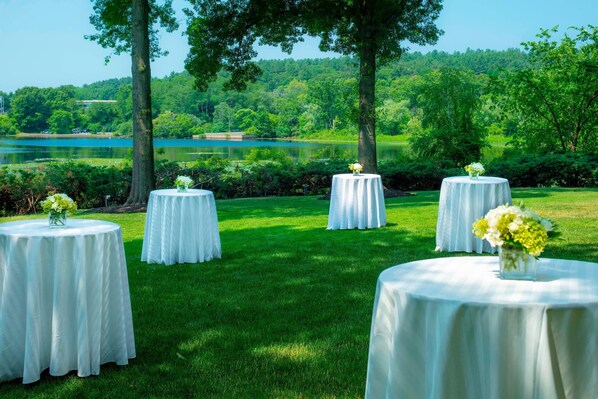 Um ambiente exterior tranquilo com mesas redondas cobertas por toalhas brancas, cada uma decorada com um vaso de flores, emoldurado por vegetação exuberante e um lago.