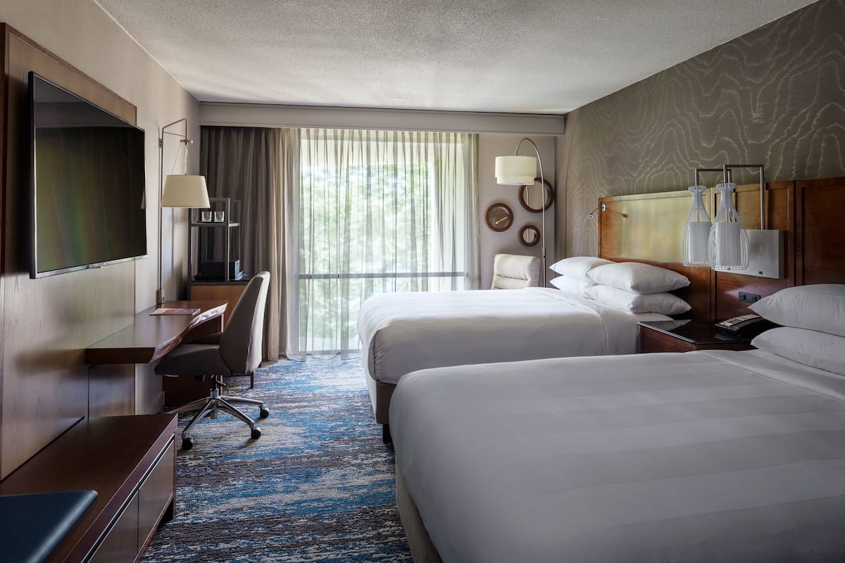 Quarto de hotel com duas camas, uma escrivaninha, uma cadeira, uma televisão e uma janela grande com cortinas.