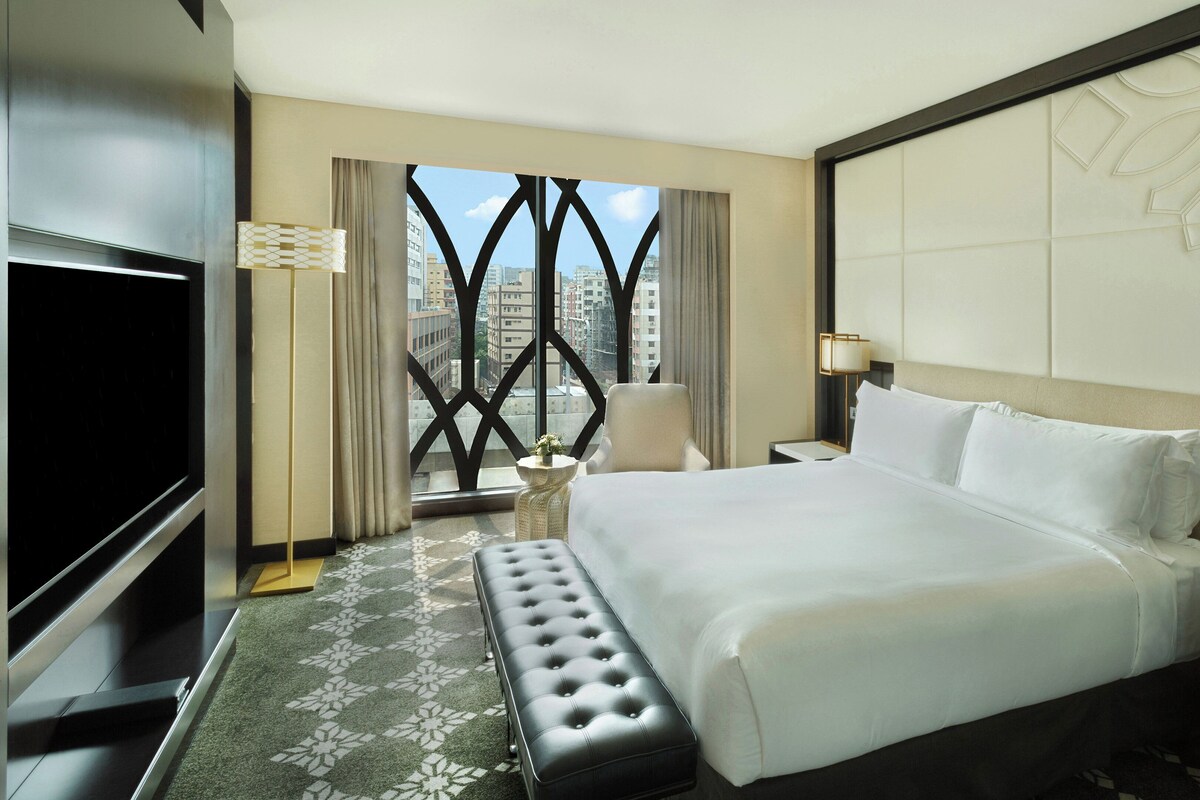 Quarto de hotel com uma cama grande, uma TV, uma escrivaninha com abajur e vista da cidade através de uma janela com design geométrico.