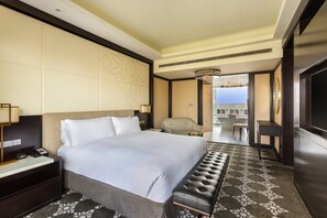 Quarto de hotel com uma cama grande, mesa de cabeceira com luminária, uma cadeira, uma mesinha e uma televisão.