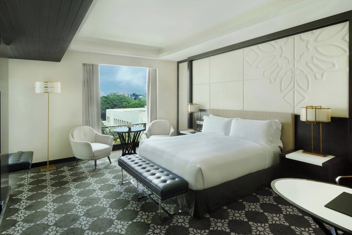 Quarto de hotel com uma cama grande, televisão de tela plana, área de estar com mesa e cadeiras, e vista para o verde lá fora.
