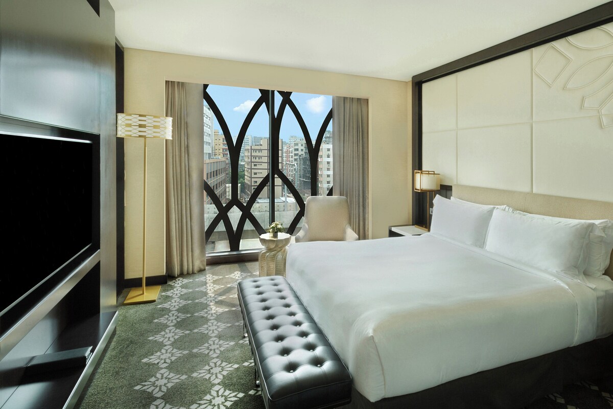 Quarto de hotel com uma cama grande, uma TV, uma escrivaninha com abajur e vista da cidade através de uma janela com design geométrico.
