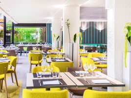 Restaurante moderno com cadeiras e mesas amarelas, grandes janelas com vista para o verde, e opções de mesas dentro e fora.