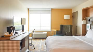 Quarto de hotel com cama, escrivaninha, cadeira e janela com persianas.