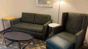 Quarto com sofá verde, um puf azul, uma pequena mesa branca e um abajur.