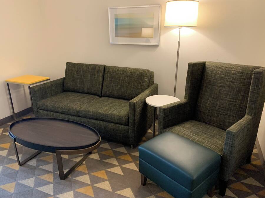 Quarto com sofá verde, um puf azul, uma pequena mesa branca e um abajur.