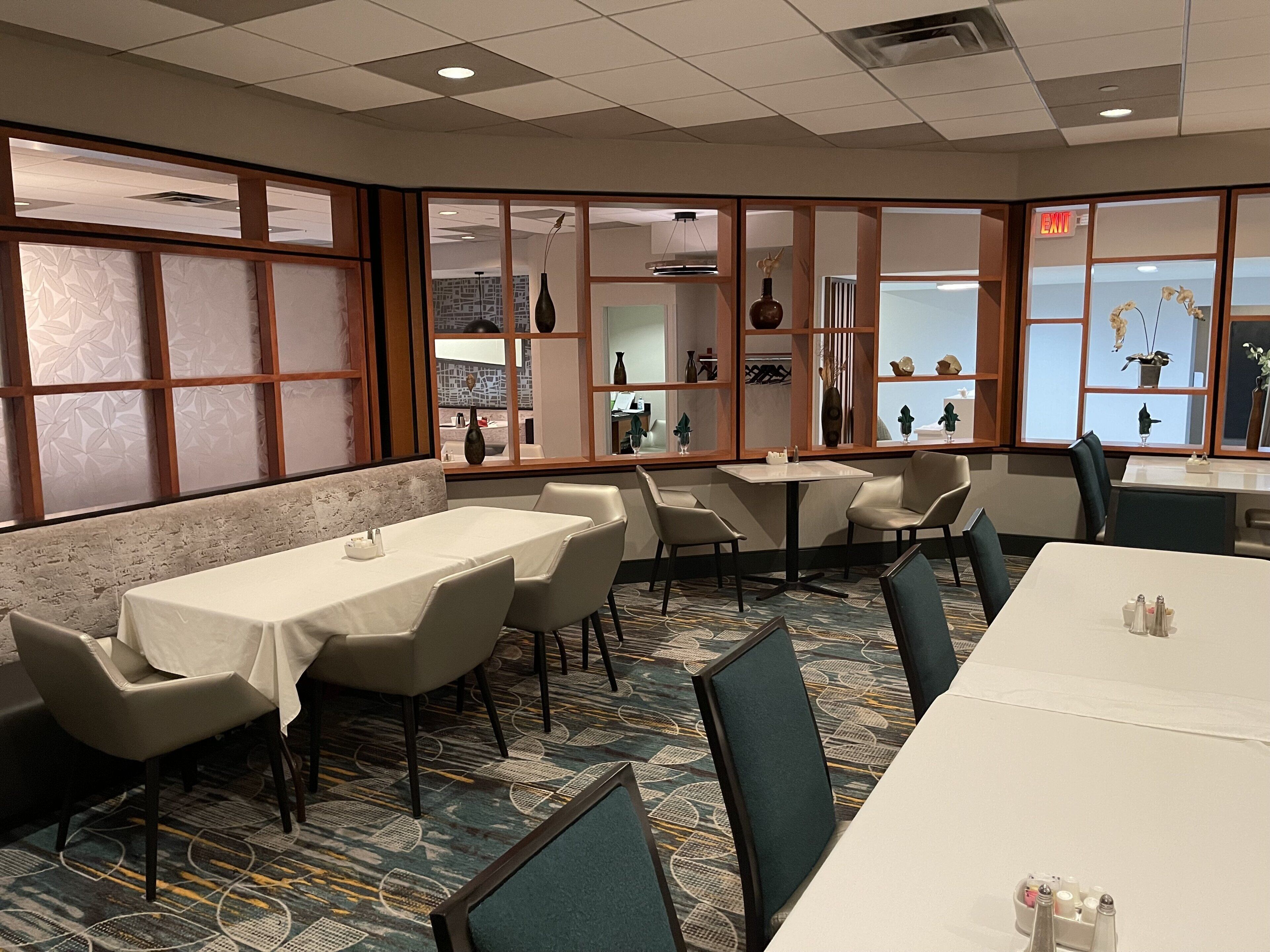 Uma sala de jantar de hotel com mesas e cadeiras, um tapete estampado e uma vista pela janela com objetos decorativos.