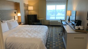 Quarto de hotel com uma cama grande, uma escrivaninha com computador, uma cadeira, uma janela com vista para árvores e um ar condicionado fixado na parede.
