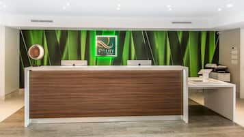 Um balcão de recepção com um fundo de cortina verde, logotipo da 'Quality Inn & Suites' e design moderno com balcão de madeira e bancada branca.