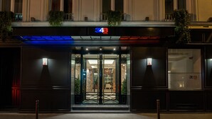 Entrada de hotel com porta de vidro, placa com a inscrição "34B HOTEL" e uma pequena planta no parapeito acima da porta.