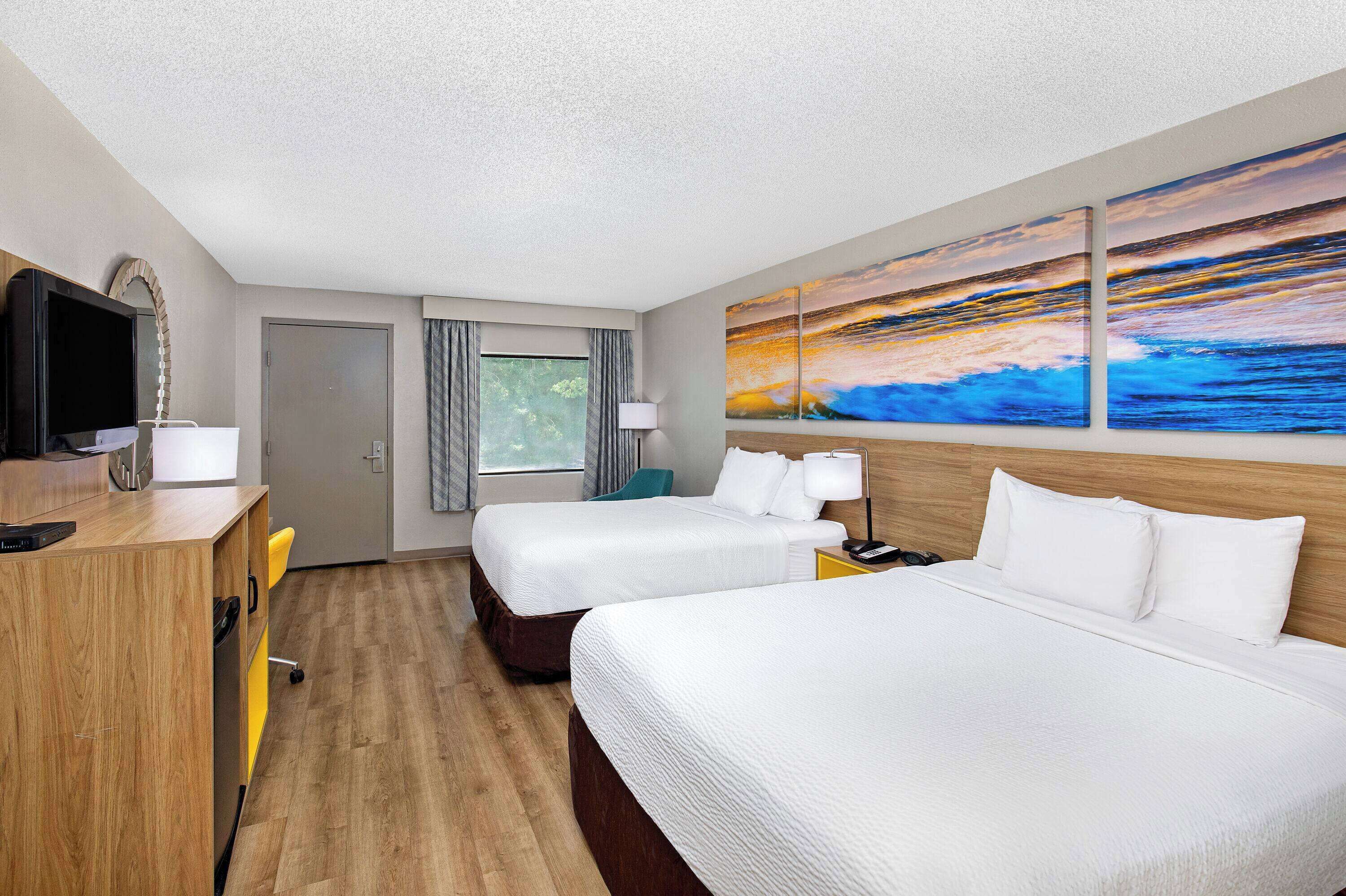 Quarto de hotel com duas camas, uma escrivaninha, televisão e uma pintura de um pôr do sol sobre o oceano.