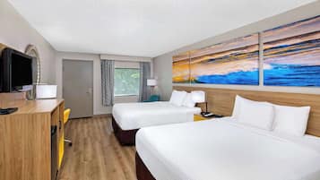 Quarto de hotel com duas camas, uma escrivaninha, televisão e uma pintura de um pôr do sol sobre o oceano.