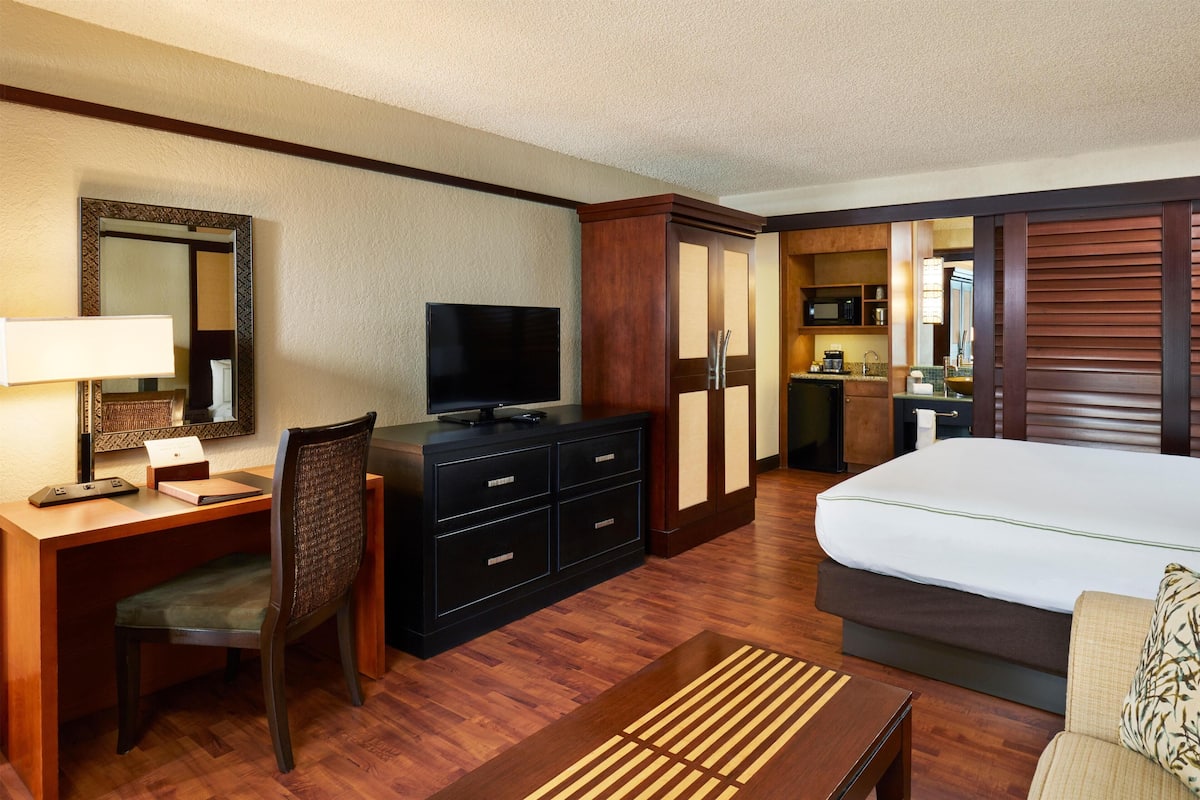 Quarto de hotel com secretária de madeira, televisão, cama, cadeira, guarda-roupa e cozinha compacta.