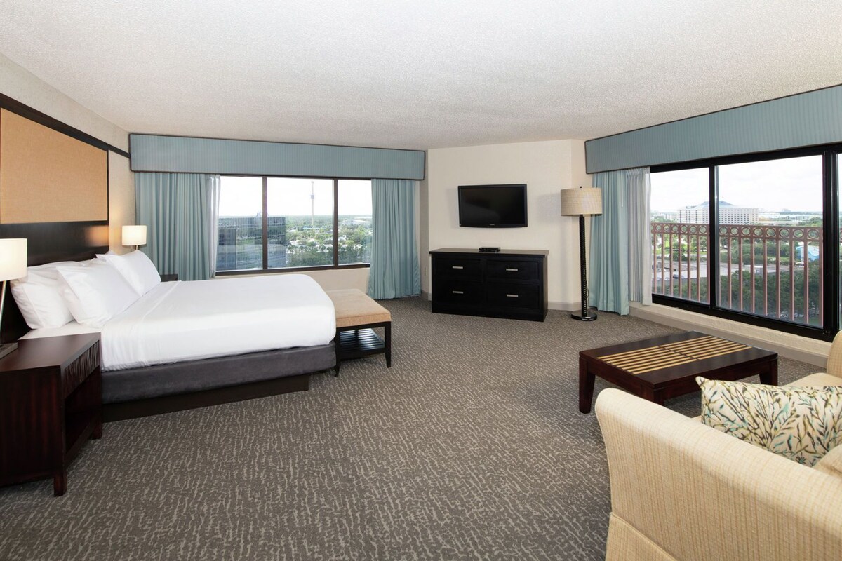 Quarto de hotel com cama, televisão, escrivaninha, cadeira e grandes janelas com vista.