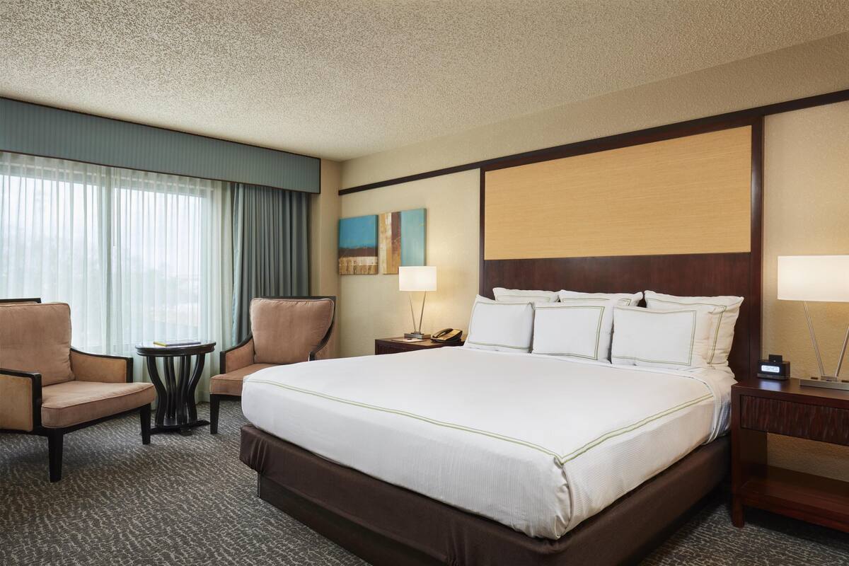 Quarto de hotel com uma cama grande, duas poltronas, uma mesinha e uma mesa de cabeceira com luminária.