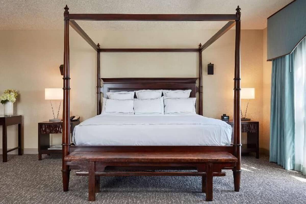 Uma cama de dossel com roupa de cama branca, duas mesas de cabeceira com luminárias, e vista das paredes e carpete bege do quarto.