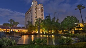 Um hotel moderno com vários andares, piscina no terraço e área externa bem iluminada com um lago.