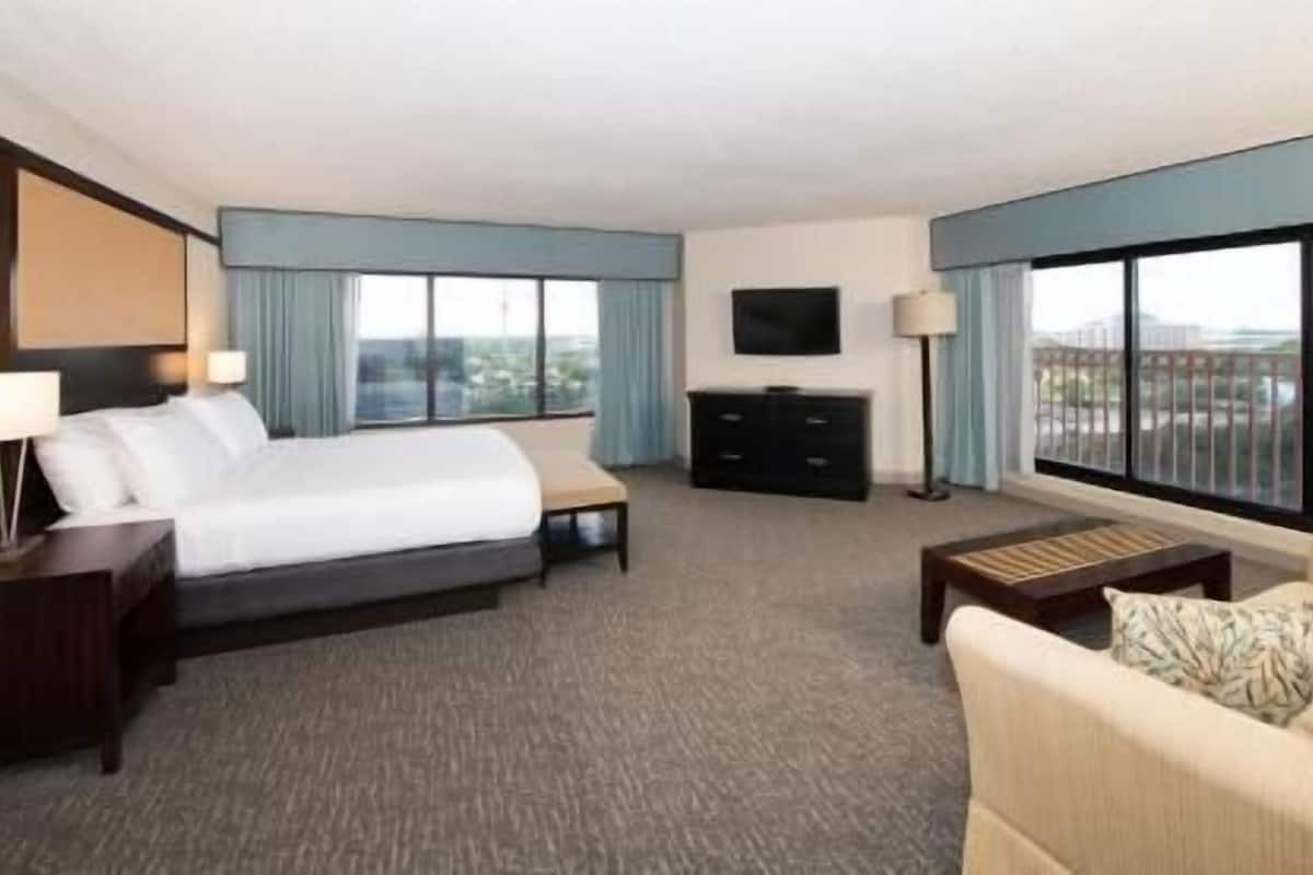 Quarto de hotel com cama, televisão, cômoda, sofá e janelas amplas.