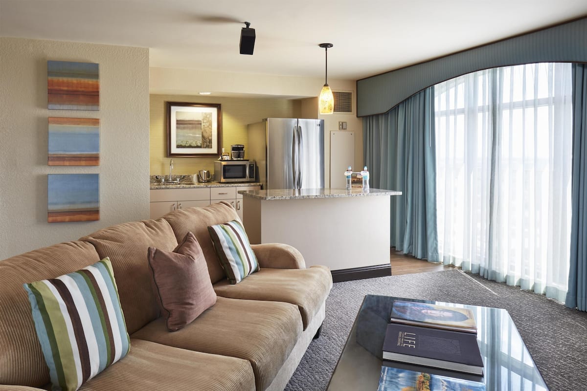 Quarto de hotel moderno com sofá, mesa de centro, cozinha e uma grande janela com cortinas.